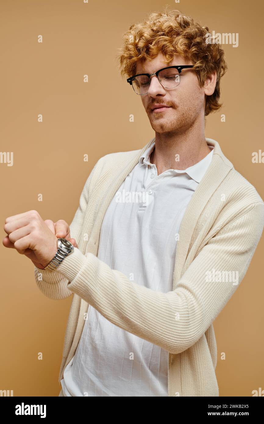 handsome young man in eyeglasses looking at wristwatch on beige ...