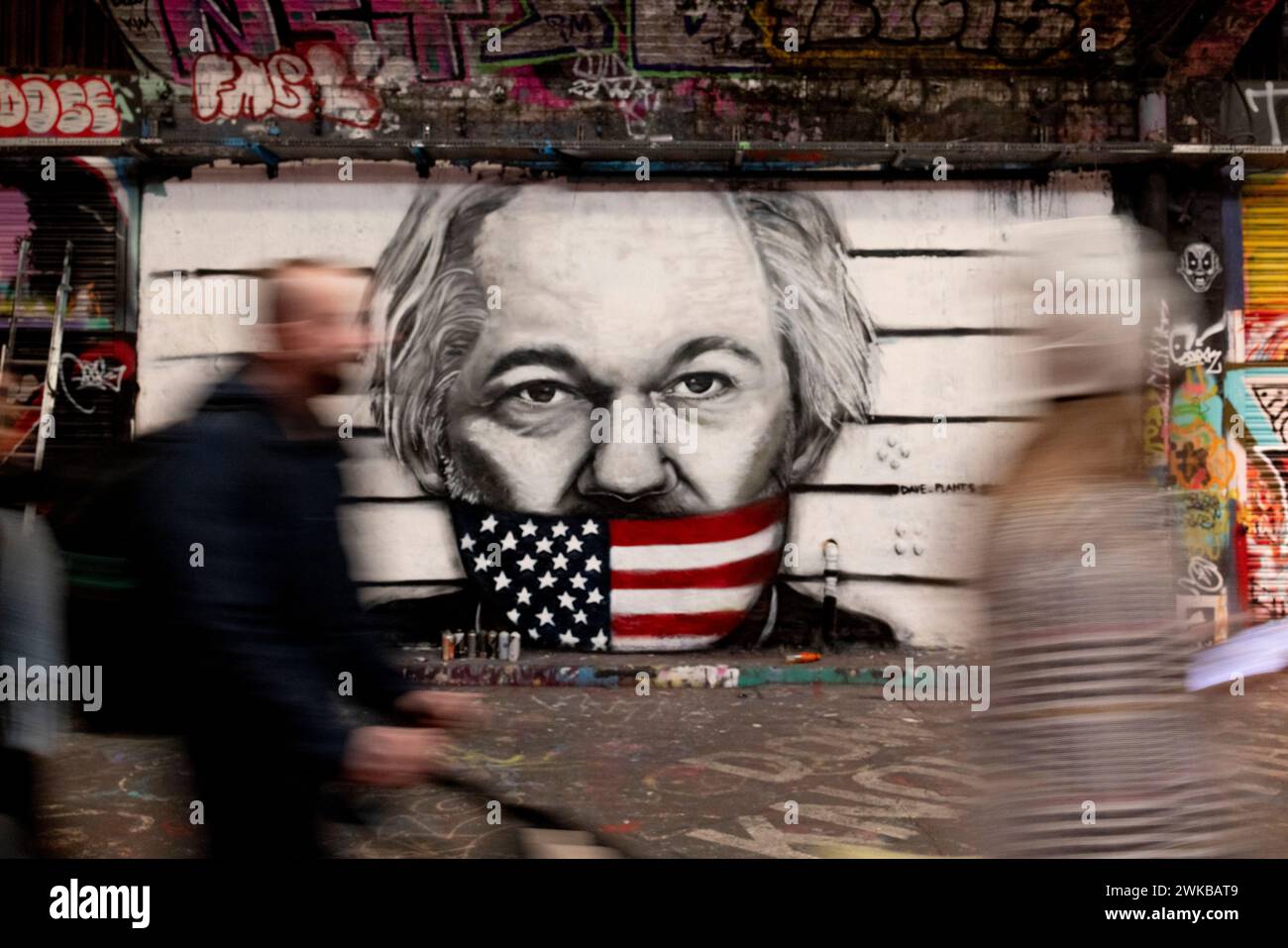 London, UK. 19th Feb, 2024. People walk by a graffiti depicting Julian ...