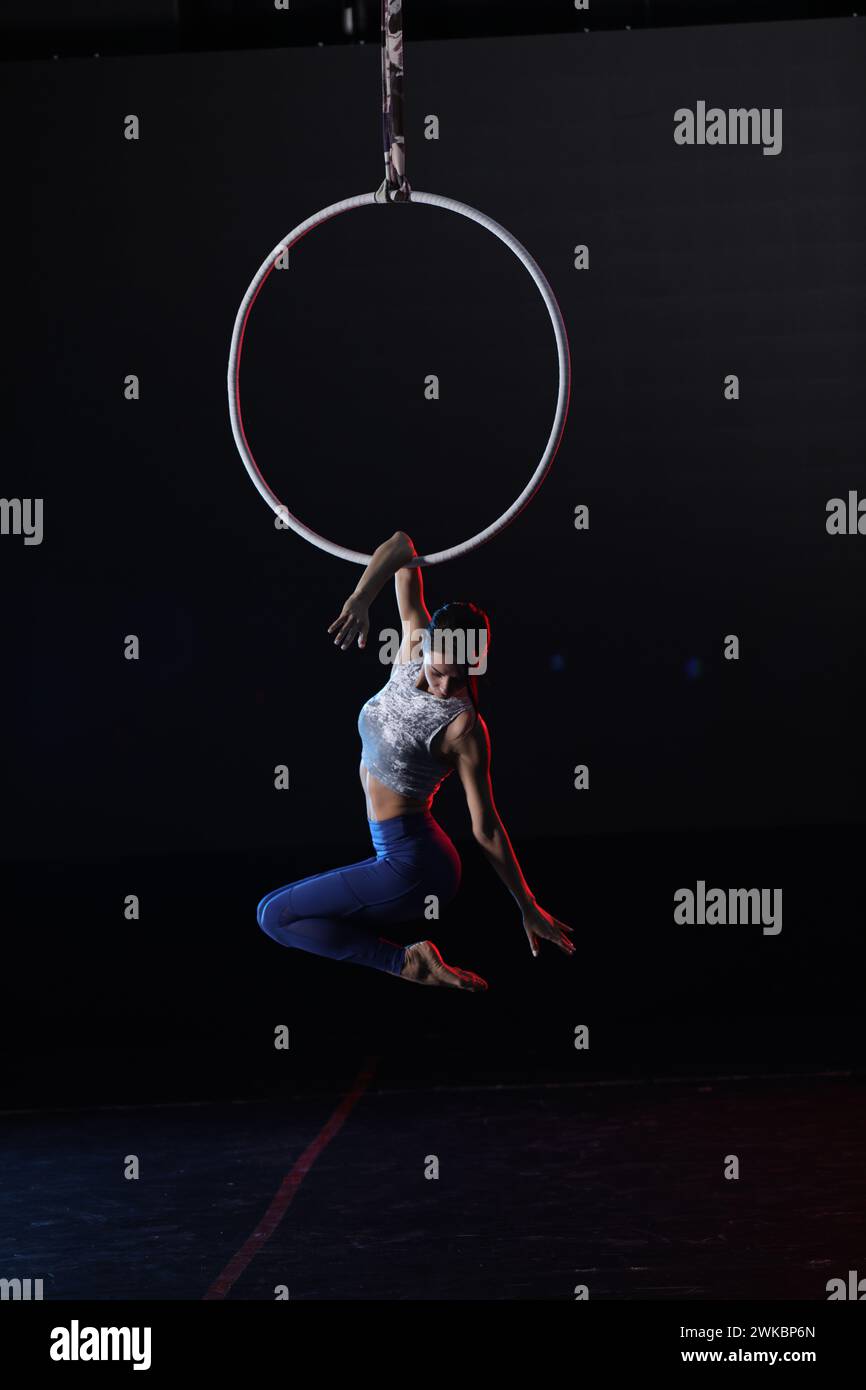 Young woman performing acrobatic element on aerial ring against dark ...