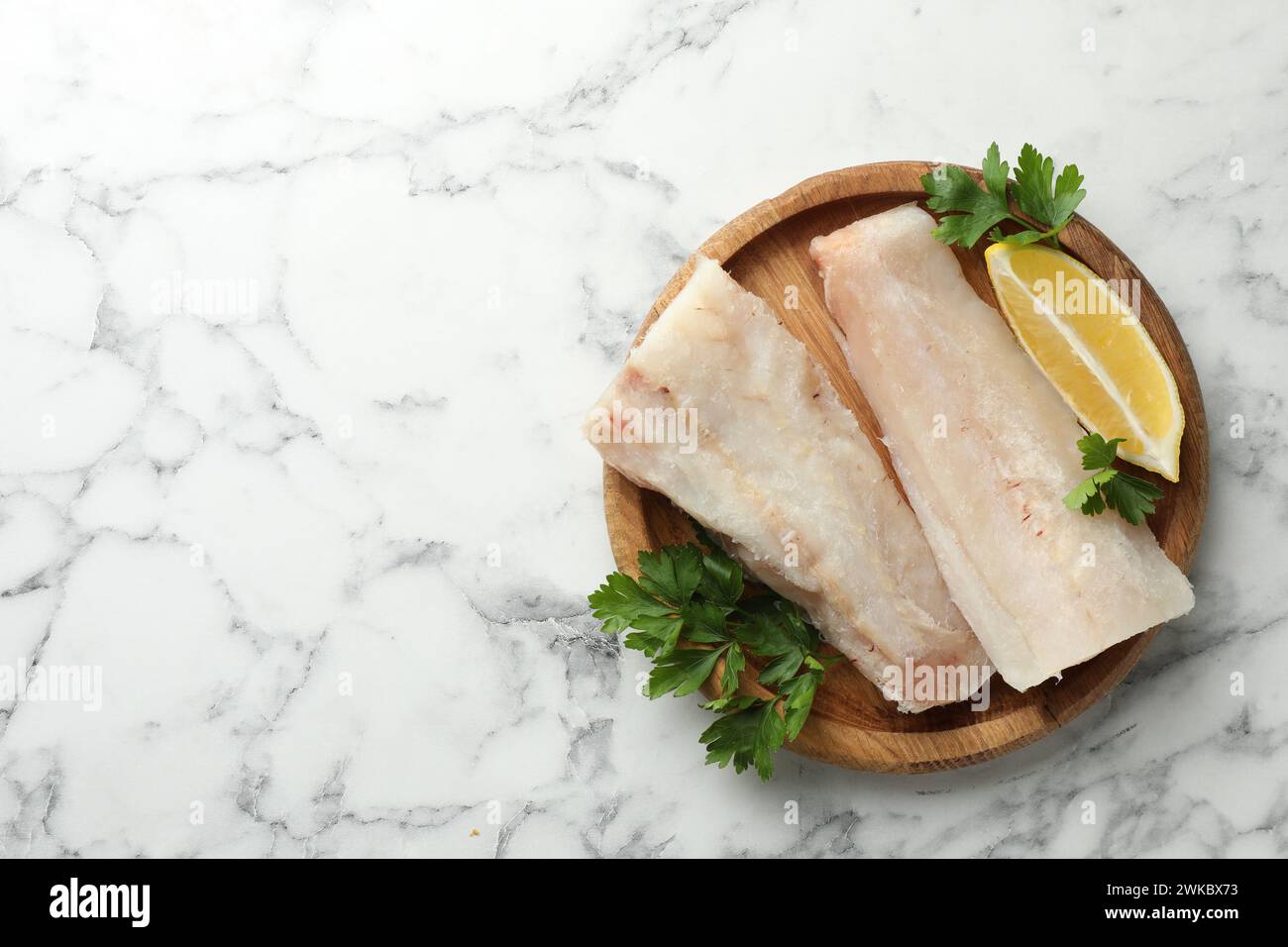 Fresh raw cod fillets with parsley and lemon on white marble table, top ...
