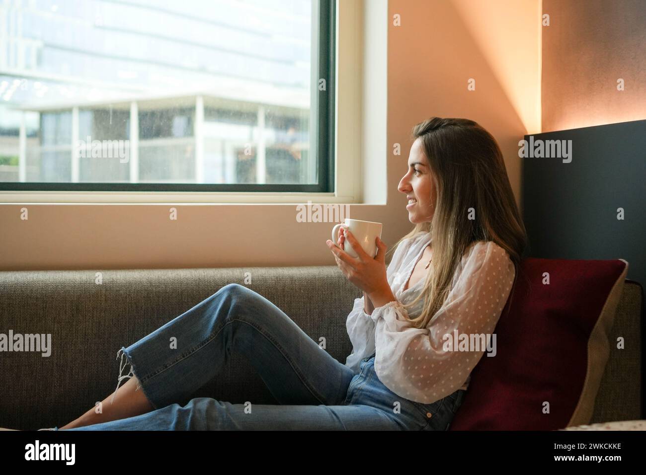 A young woman sits comfortably by the window of a hotel room, savoring ...