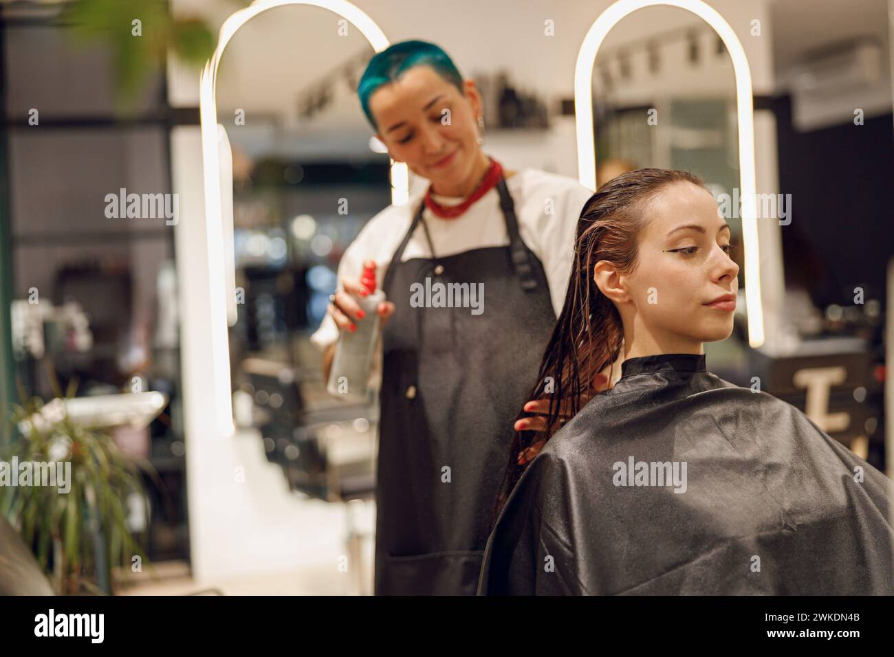 Stylish female hairdresser applying spray on wet hair to woman in ...