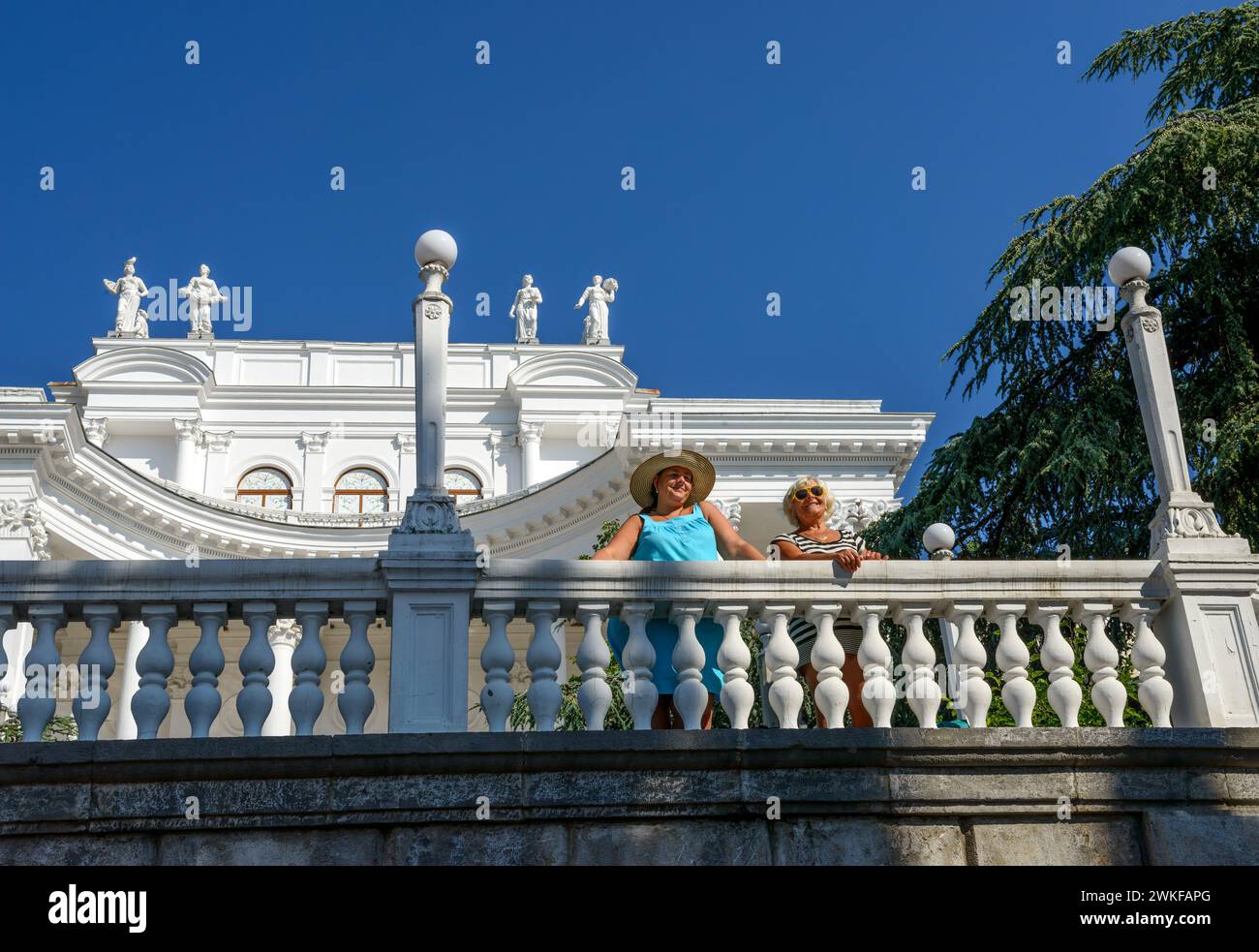 Two women are posing near balustrade in park with white building of ...