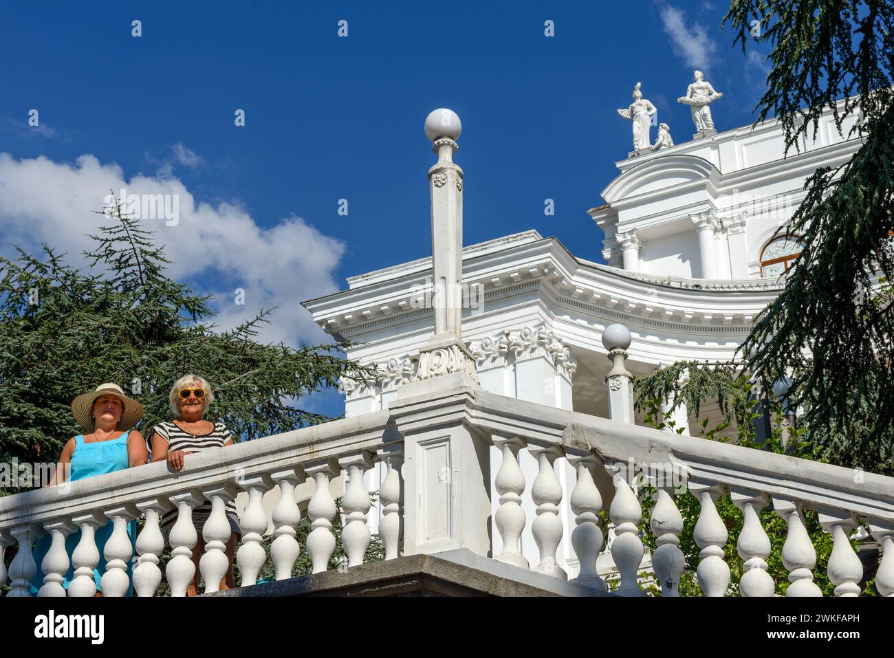 Two women are posing near balustrade in park with white building of ...