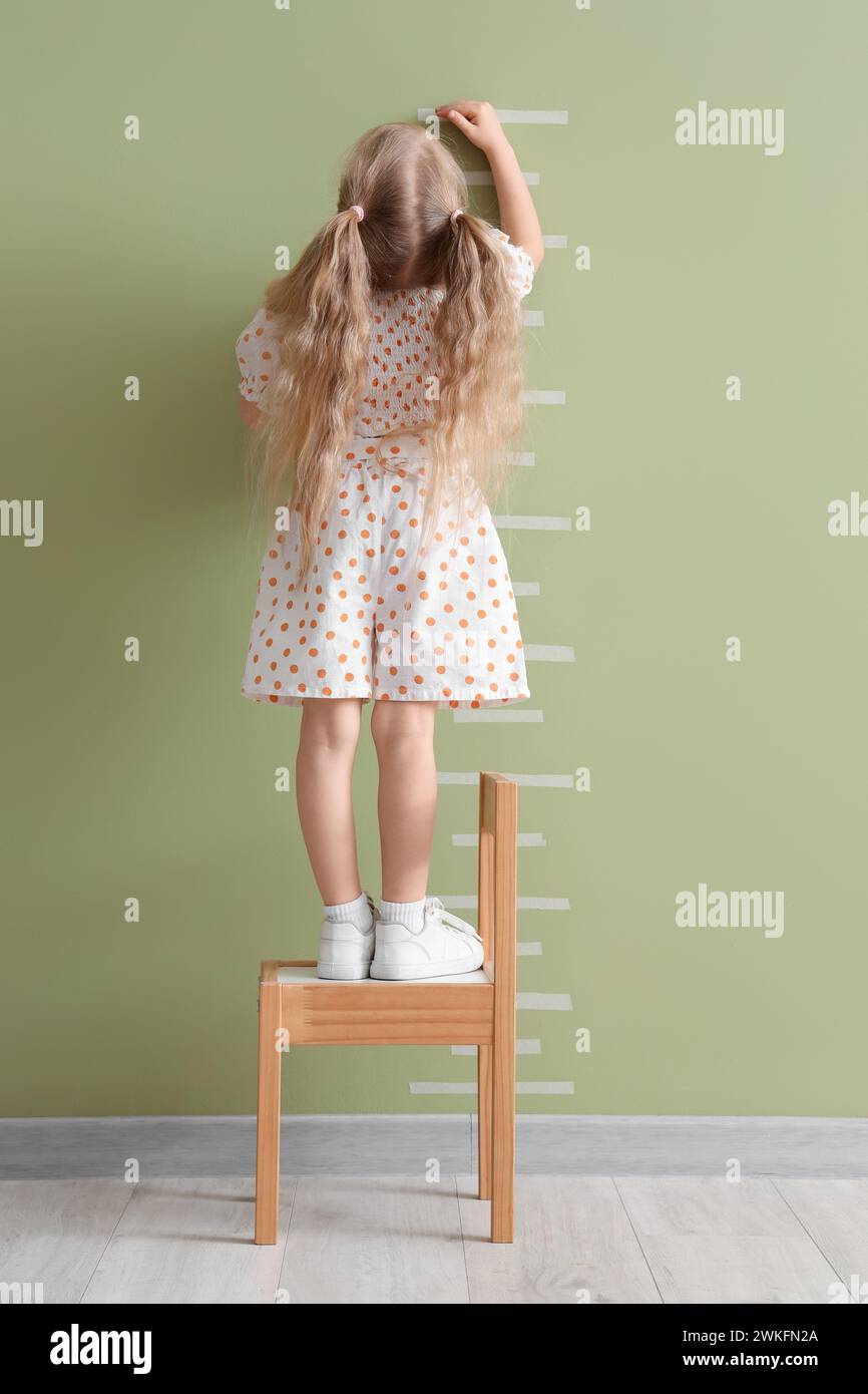 Cute little girl standing on chair and measuring height near green wall ...