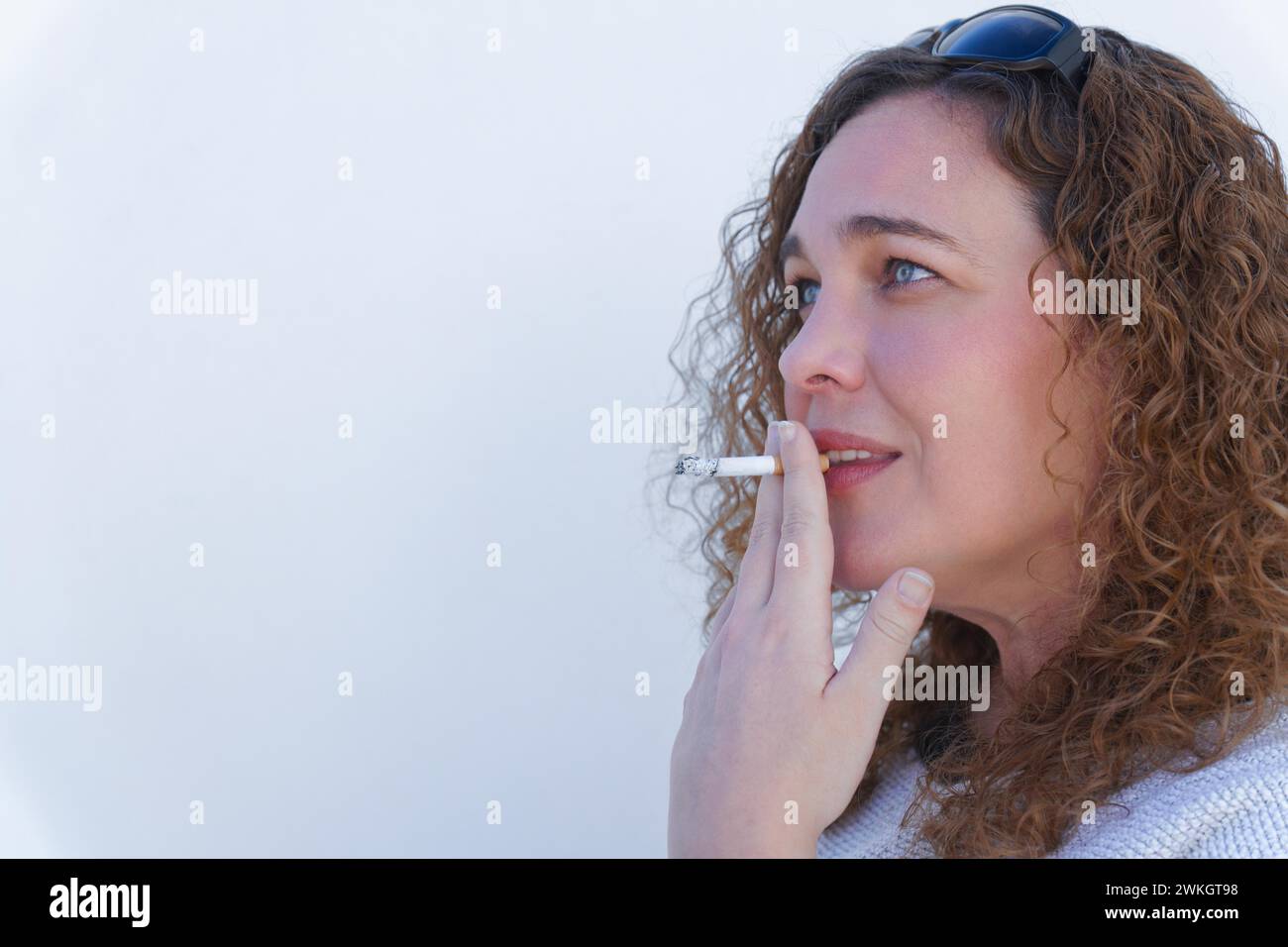 Attractive blue-eyed, curly-haired woman in profile smoking a cigarette with white background ...