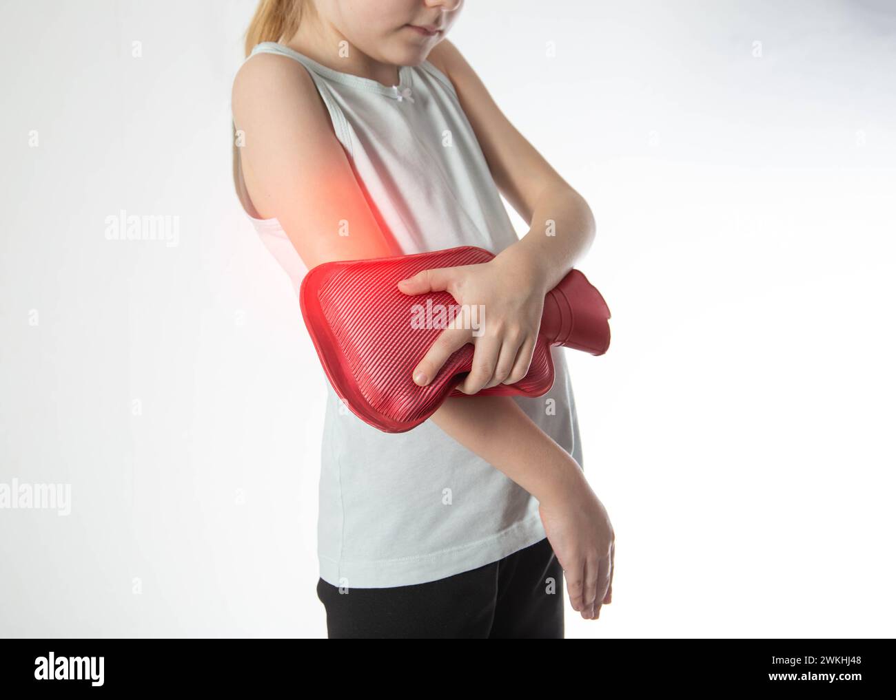 A little girl applies a red heating pad with hot water to her elbow ...