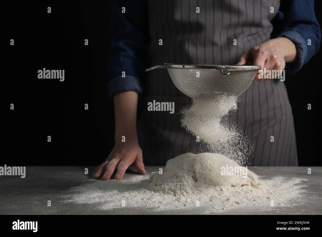 Making bread. Woman sifting flour over dough at table on dark ...