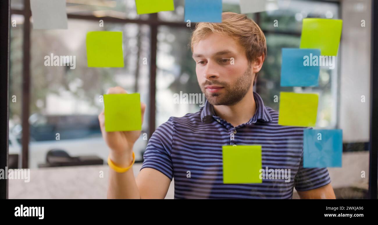 Young businessman standing near glass window with sticky notes. Man ...