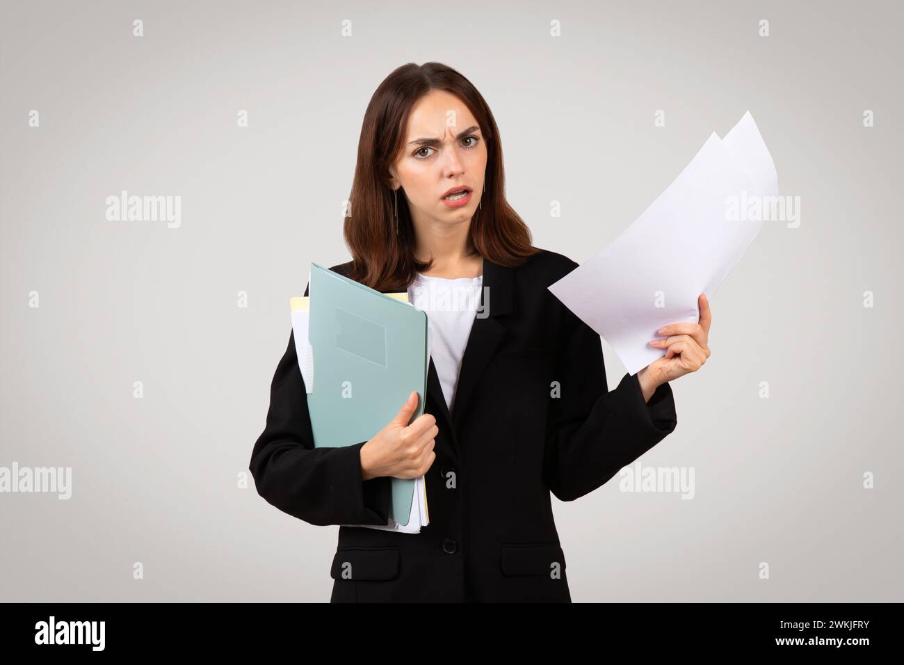 A puzzled businesswoman in a suit frowns while holding documents and a ...