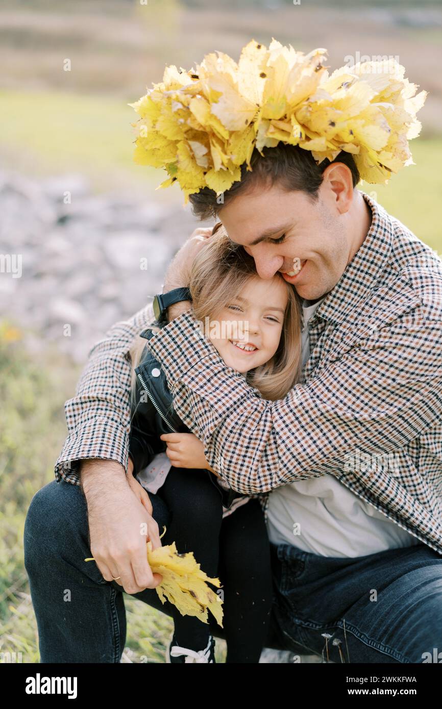 Smiling dad in a wreath of yellow leaves hugs a little girl sitting on ...