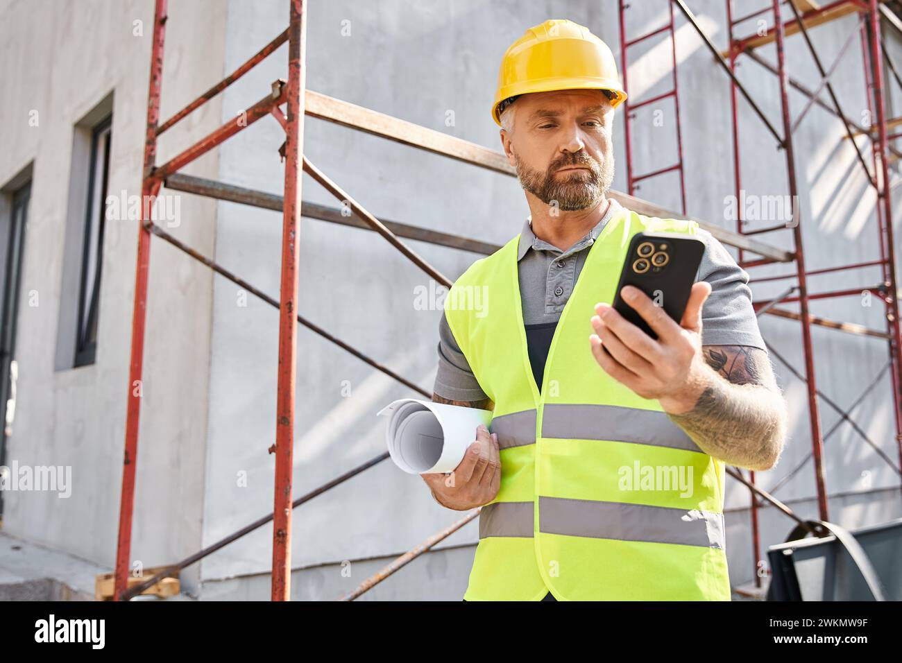 handsome bearded construction worker with tattoos and blueprint looking at phone, cottage ...