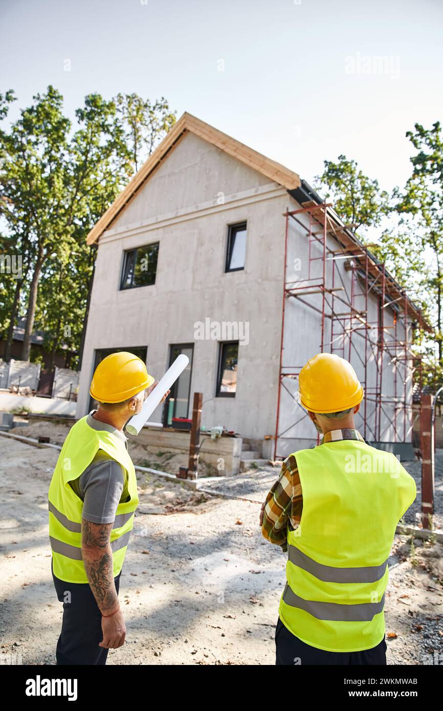 back view of devoted cottage builders in safety helmets with blueprint ...