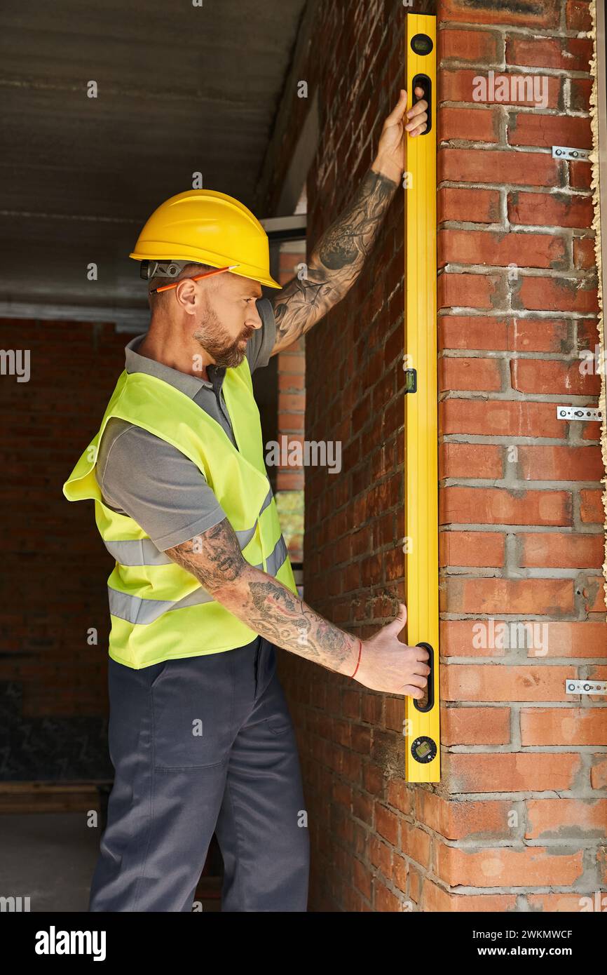 handsome bearded construction worker in safety vest measuring brick wall with level, builder ...