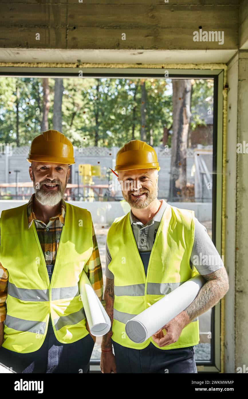 cheerful dedicated builders in safety vests and helmets looking at camera on construction site ...