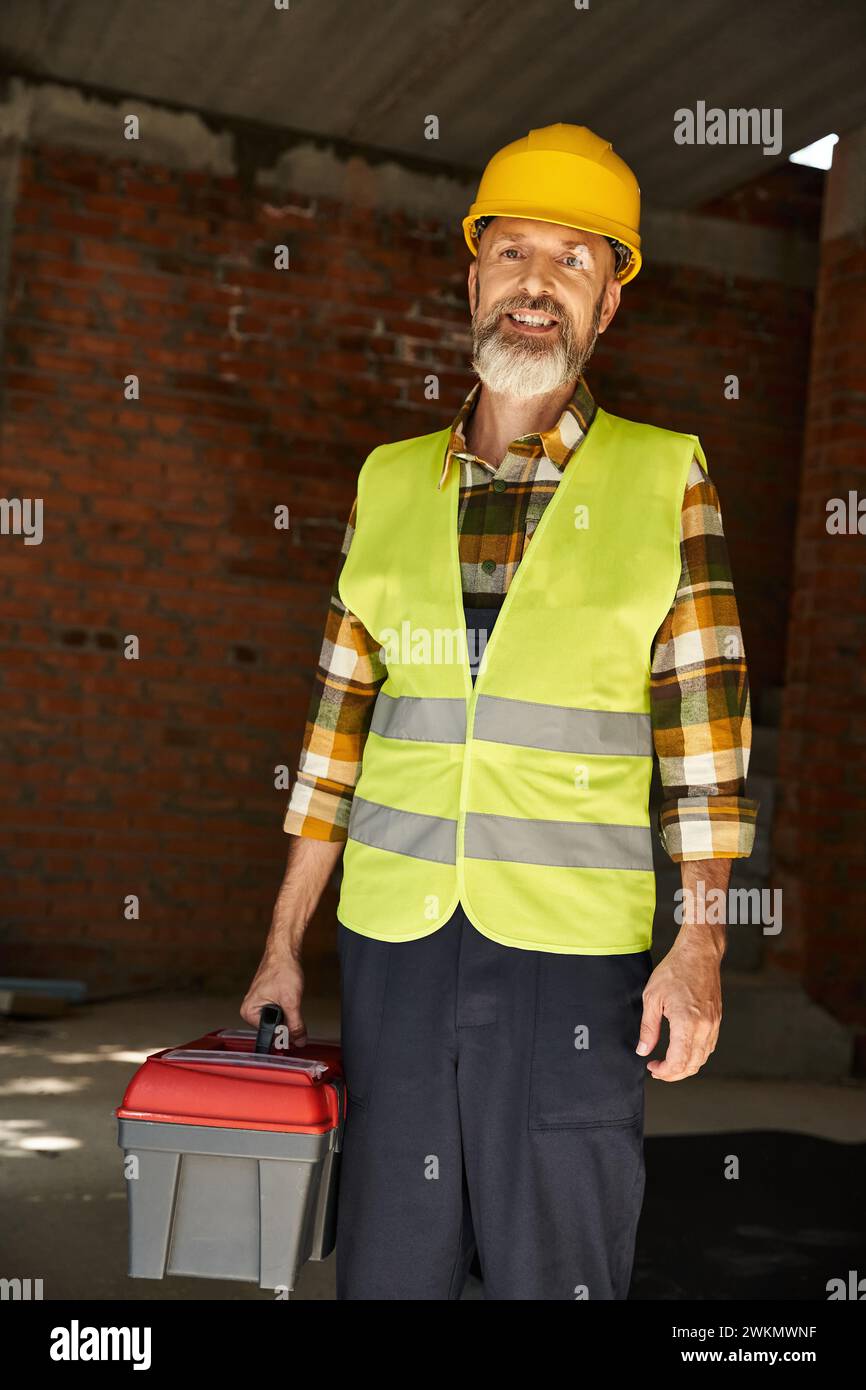 cheerful good looking builder in safety vest and helmet holding toolbox ...