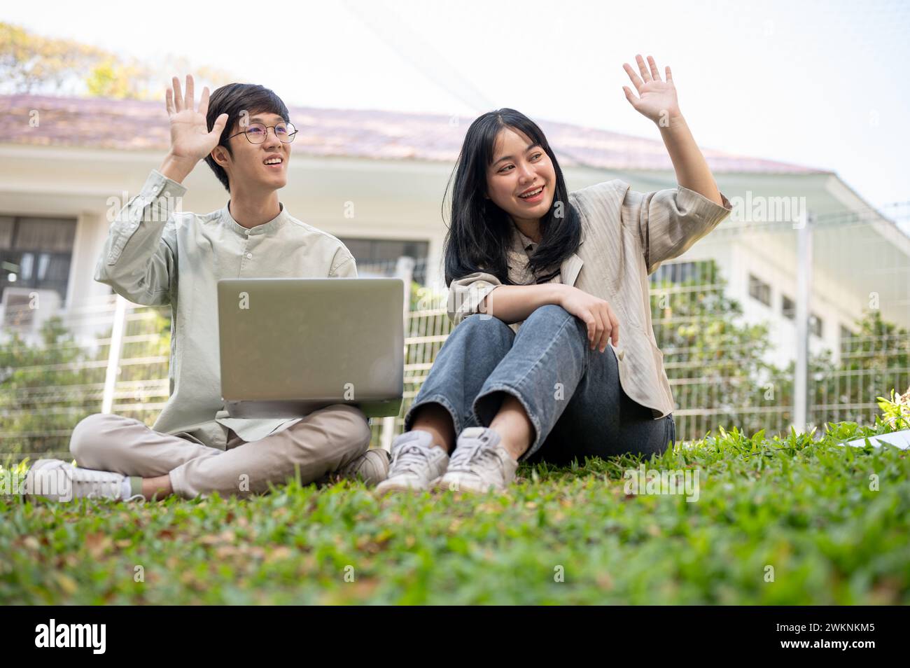 Two cheerful and friendly Asian university students are sitting on the grass in a campus park ...