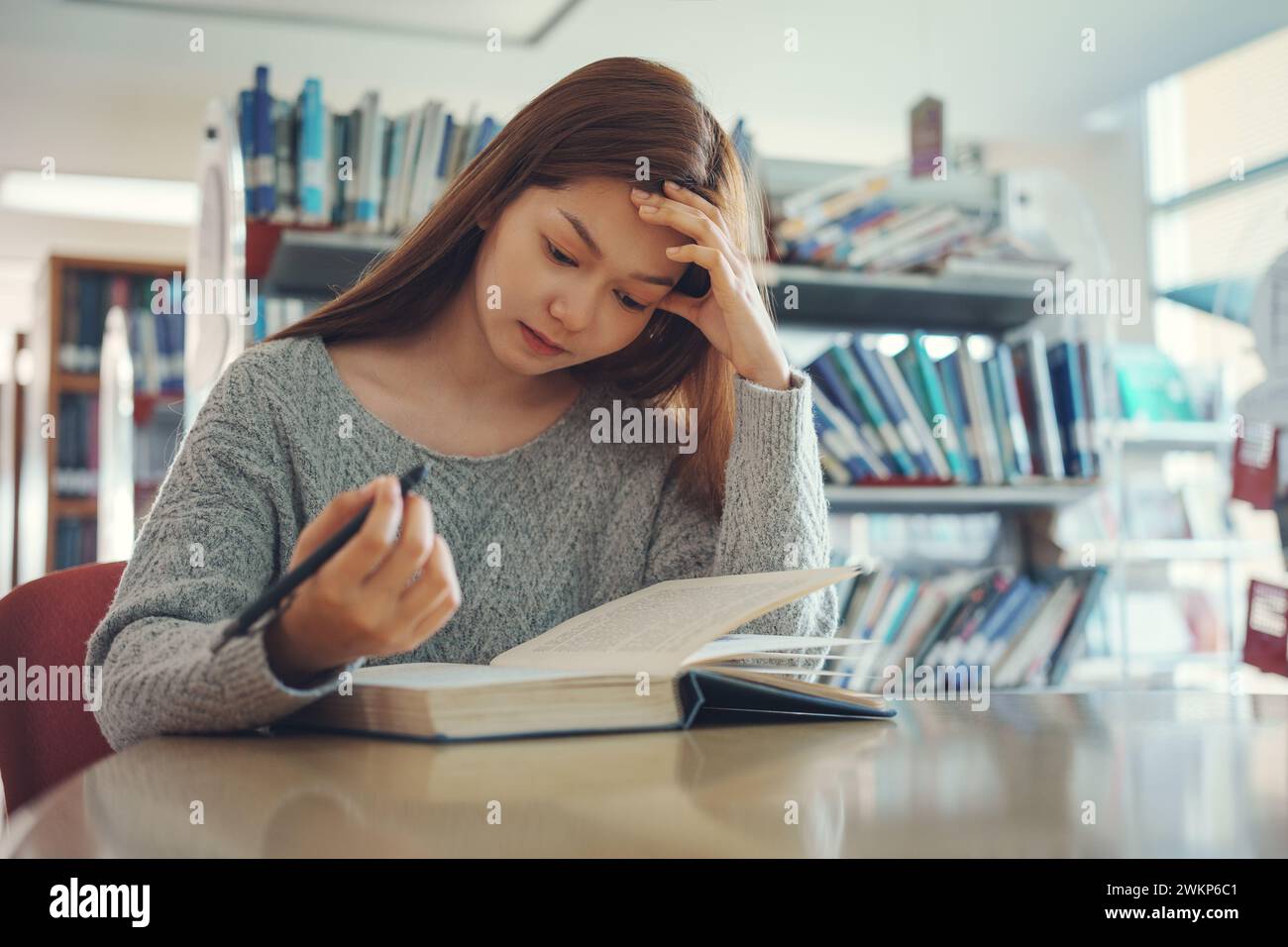 Stressed young female student sitting at wooden table and reading book ...