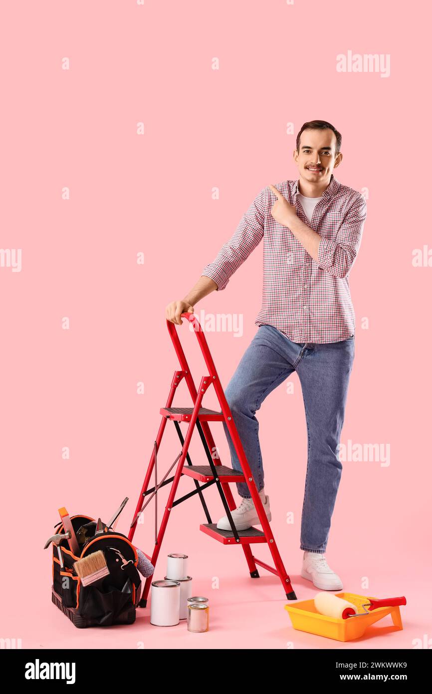Young man with ladder and builder's tools pointing at something on pink background Stock Photo ...