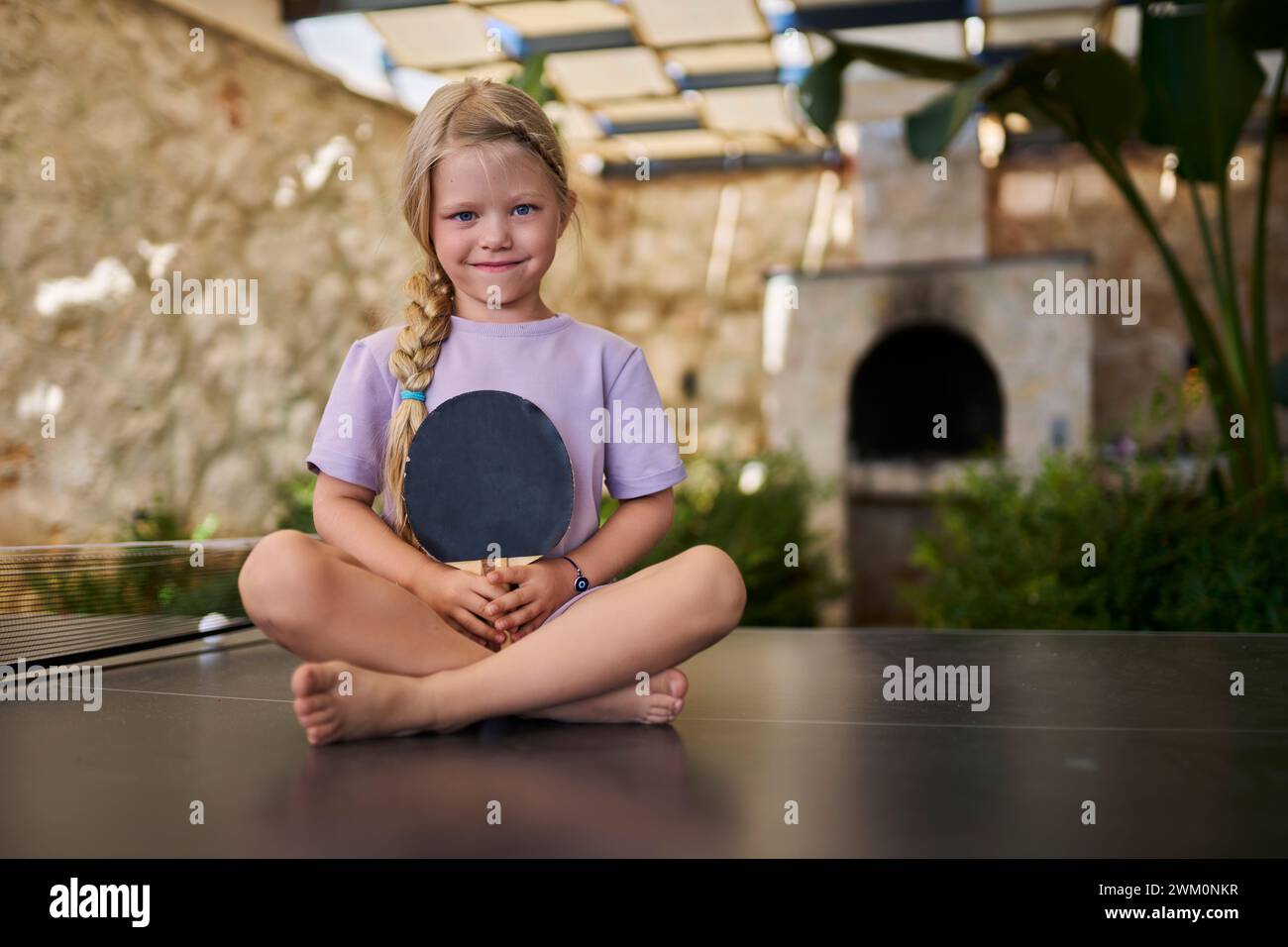 Smiling girl holding table tennis racket sitting cross-legged at villa ...