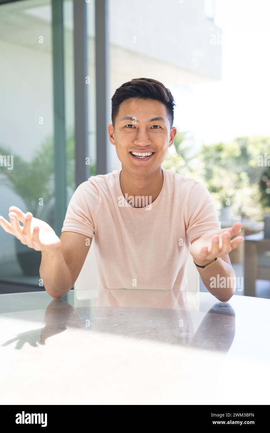 Young Asian man gestures while sitting at a table on a video call. His ...