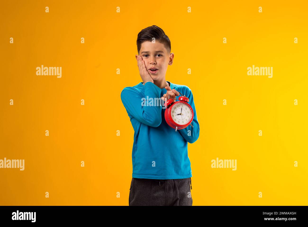Portrait of surprised child boy holding alarm clock over yellow ...
