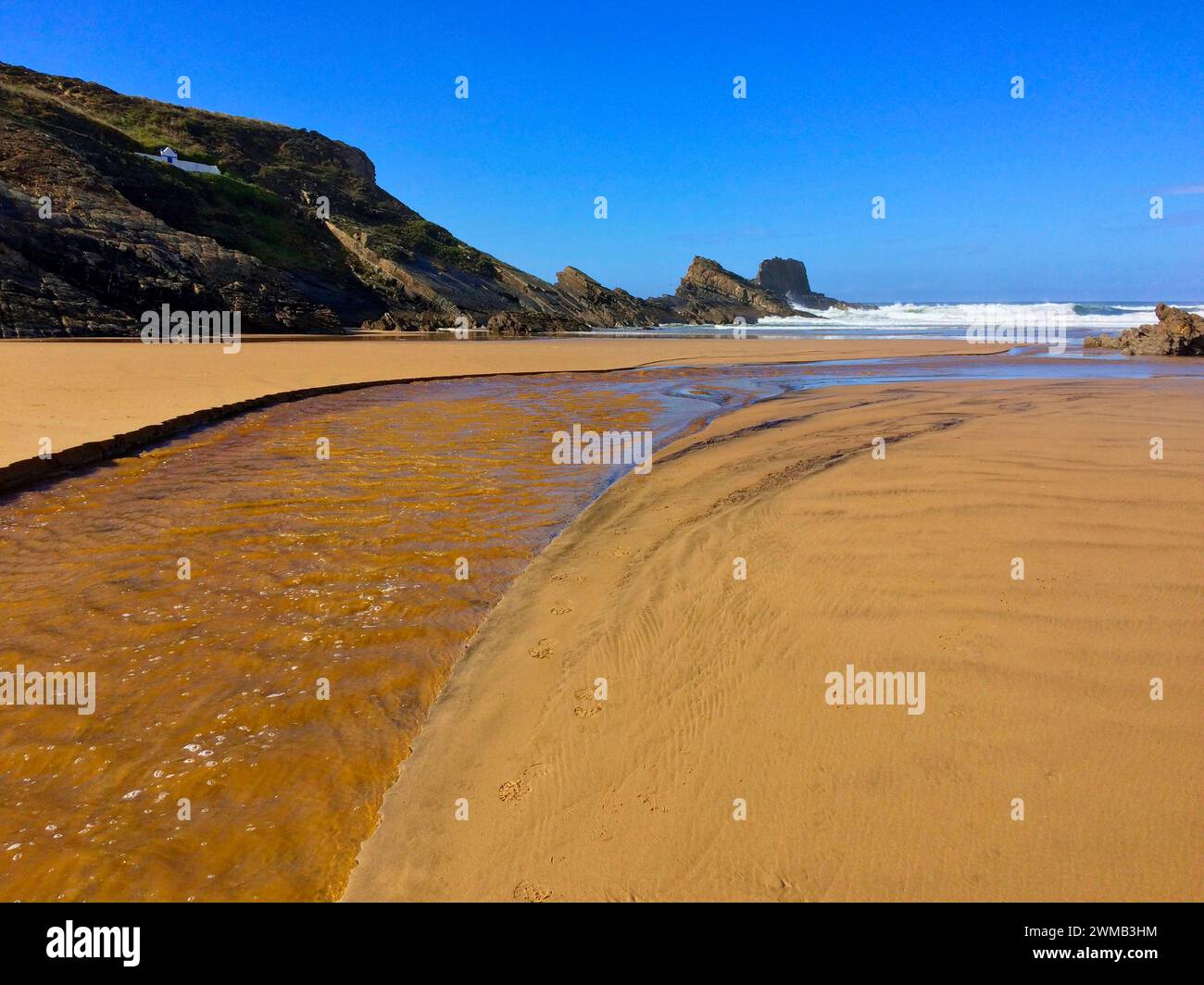 A sandy beach with a stream, surrounded by rocky cliffs under a clear ...