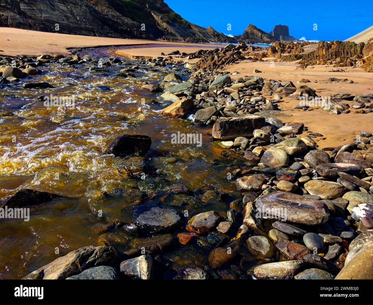 A rocky stream flows into a sandy beach, surrounded by cliffs under a ...