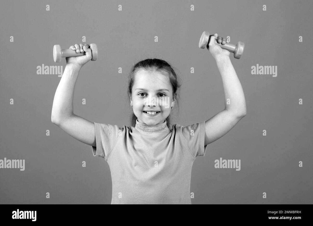 Cute little girl doing exercises with dumbbells in yellow background ...