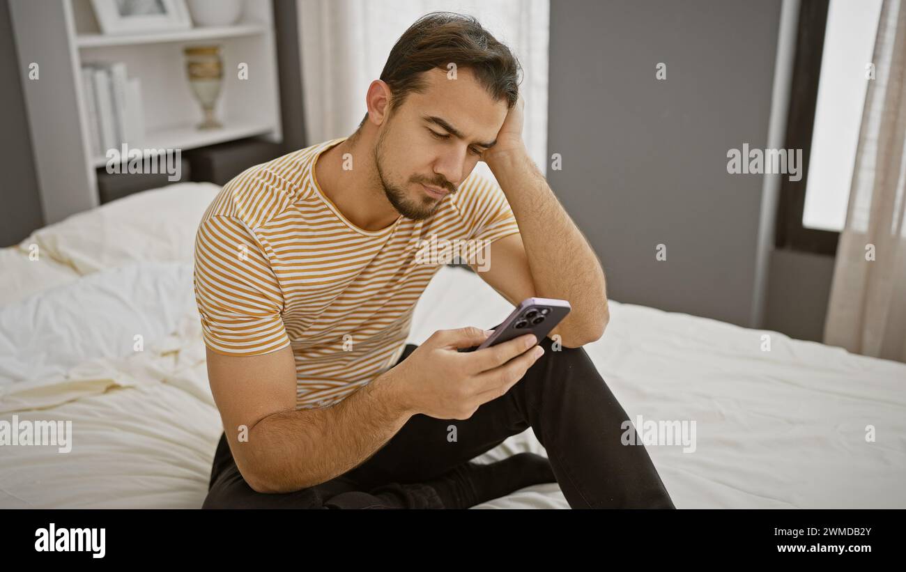 A handsome young hispanic man with a beard in casual clothing sits thoughtfully on the bed in a ...
