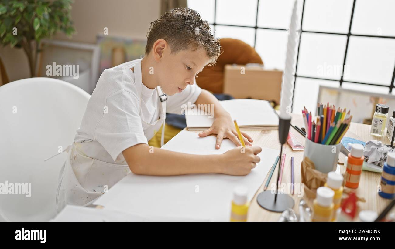 Adorable little blond boy artist intently drawing in his notebook ...