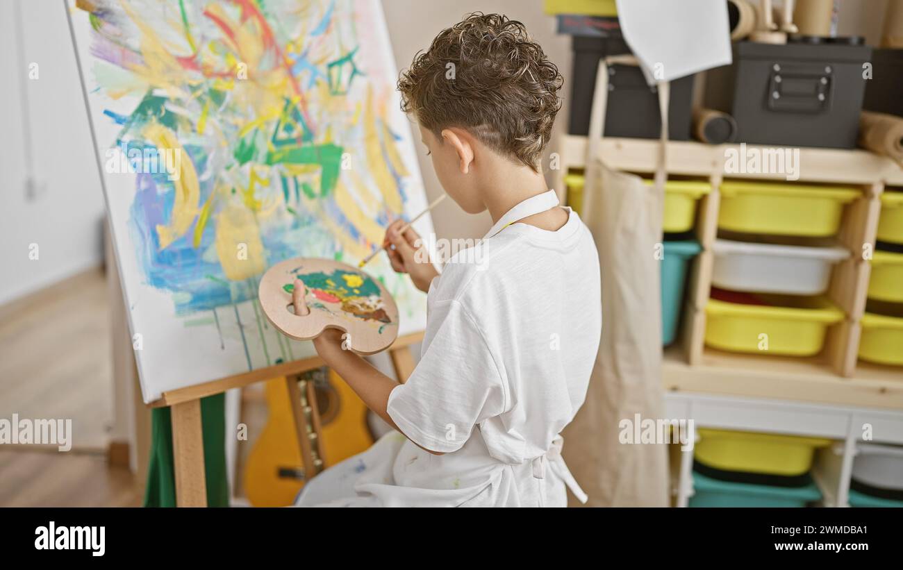 Adorable blond boy artist confidently drawing on canvas indoors, a ...