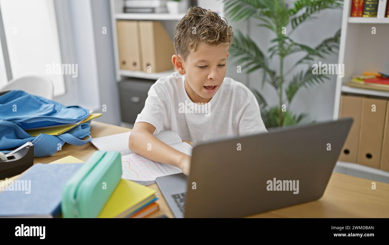 Adorable blond boy student intently focused on studying with laptop in ...