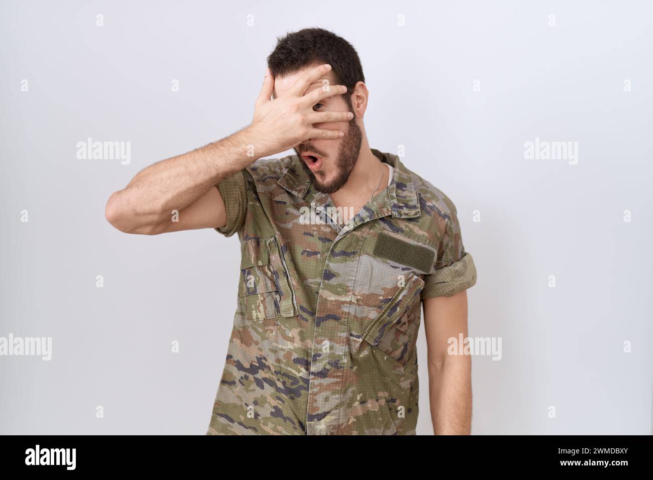 Young hispanic man wearing camouflage army uniform peeking in shock ...