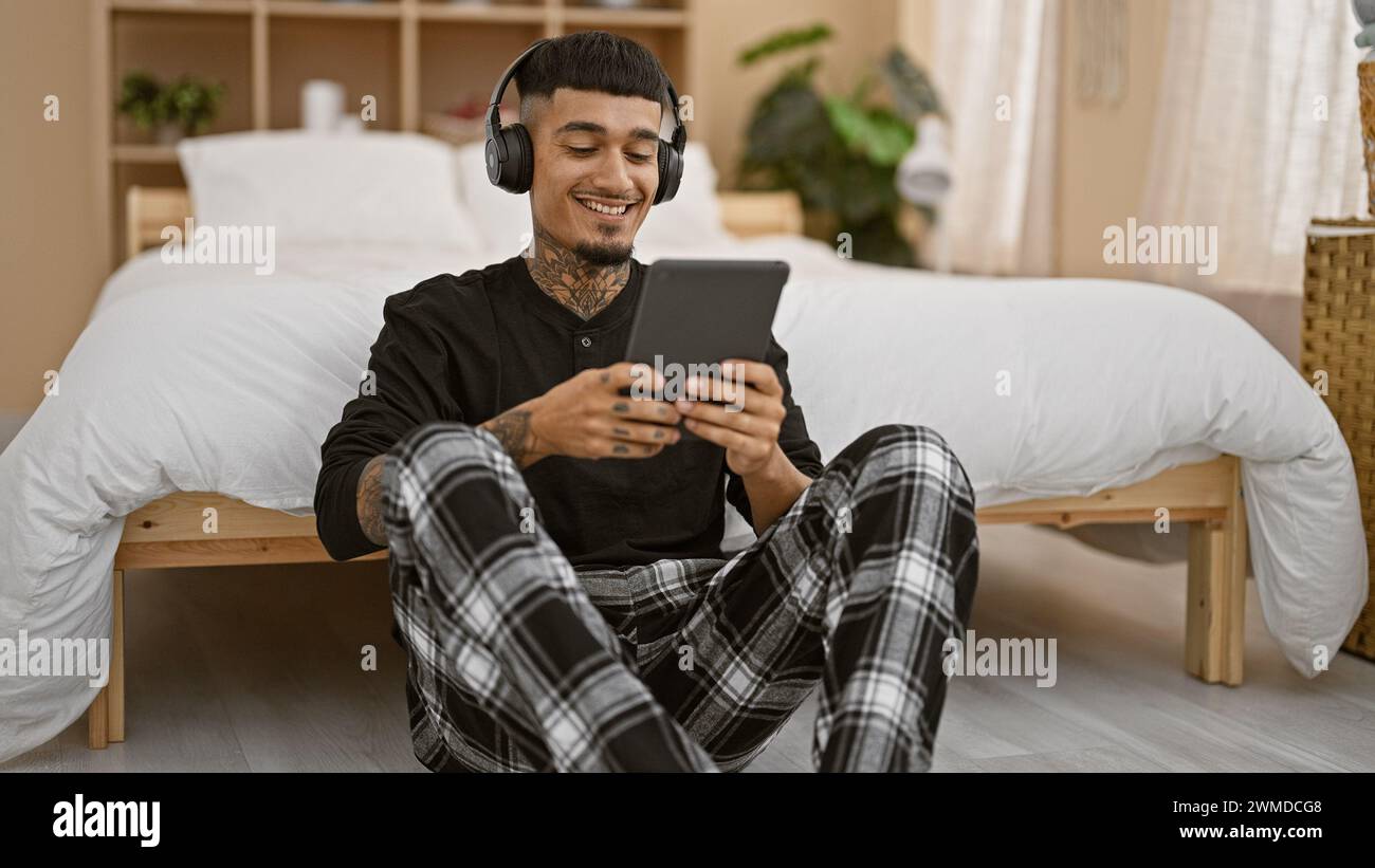 Young latin man using touchpad sitting on the floor wearing headphones at bedroom Stock Photo ...