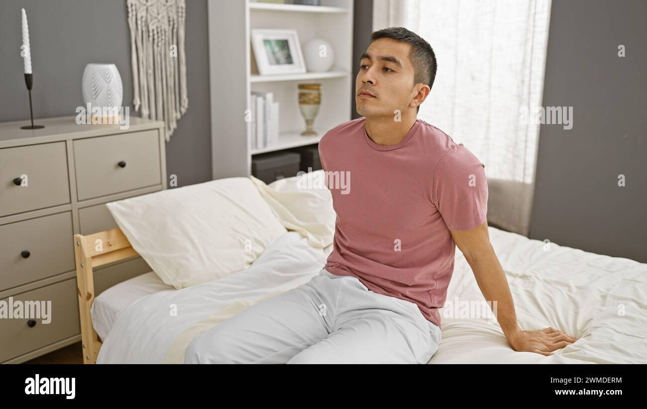 Handsome young hispanic man sitting thoughtfully on a bed in a modern bedroom interior Stock ...