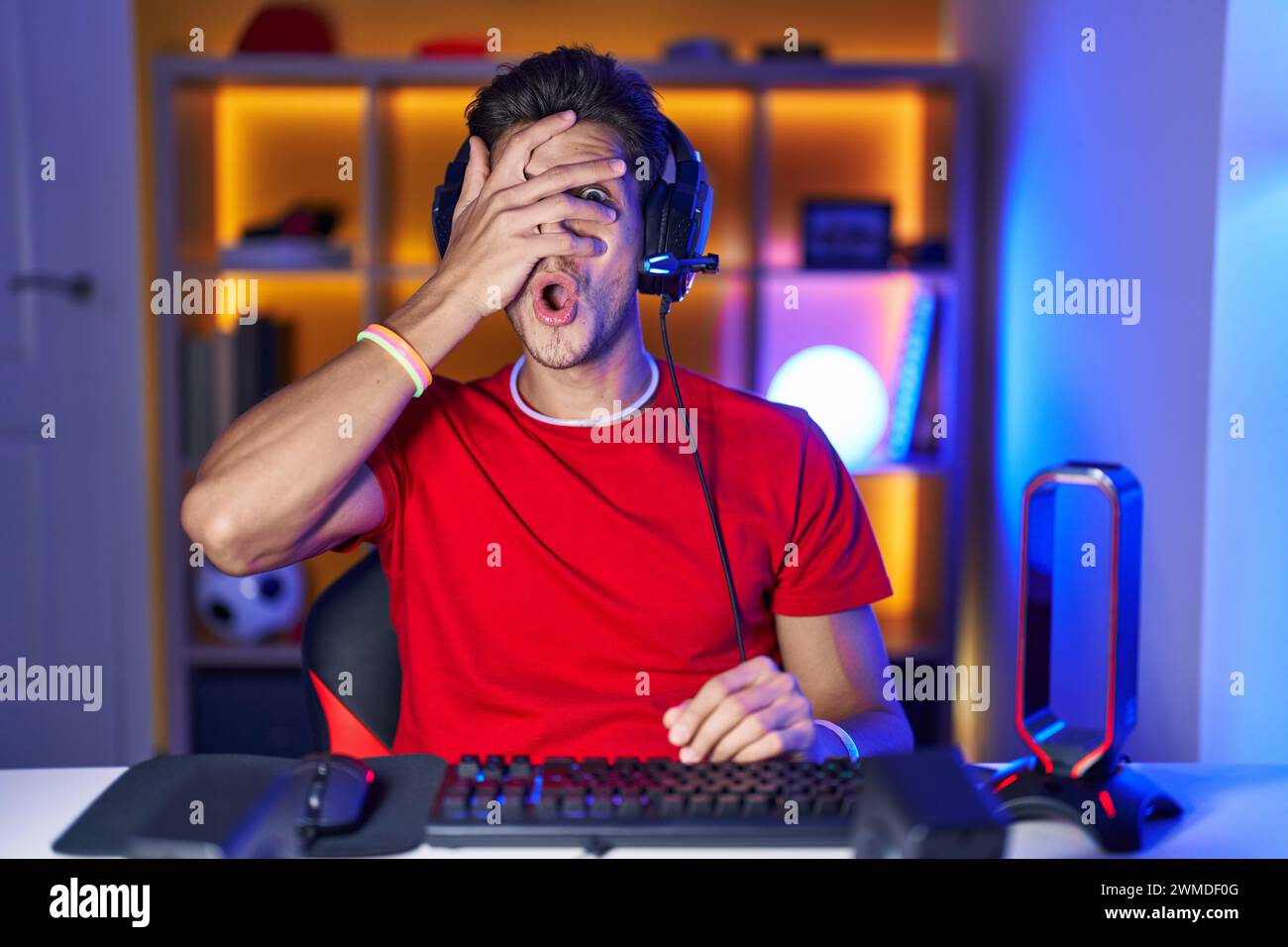 Young hispanic man playing video games peeking in shock covering face ...