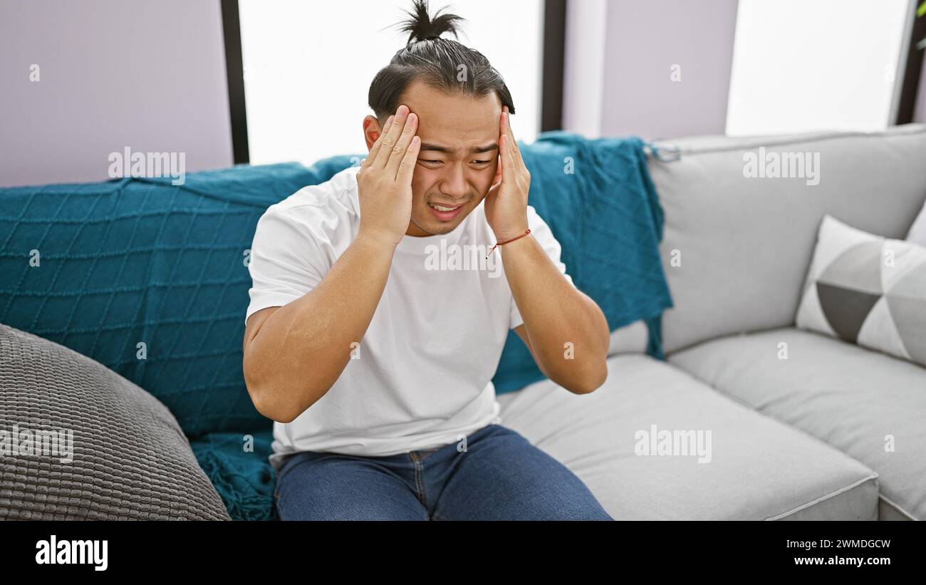 Stressed young chinese man, frustrated and alone, sits on sofa nursing ...