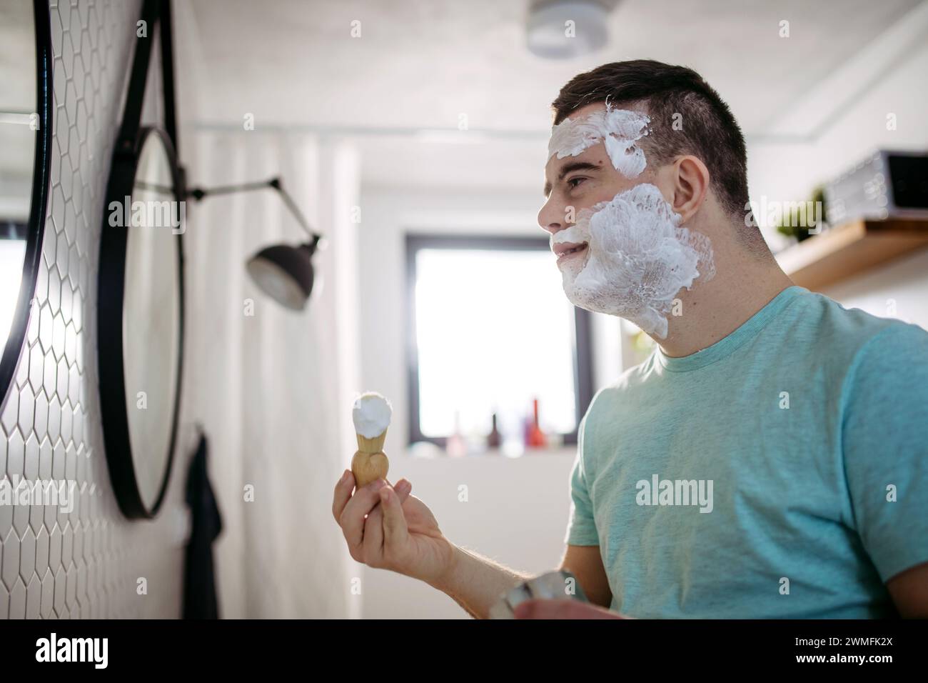 Young man with down syndrome learning how to shave, applying shaving foam all over his face ...