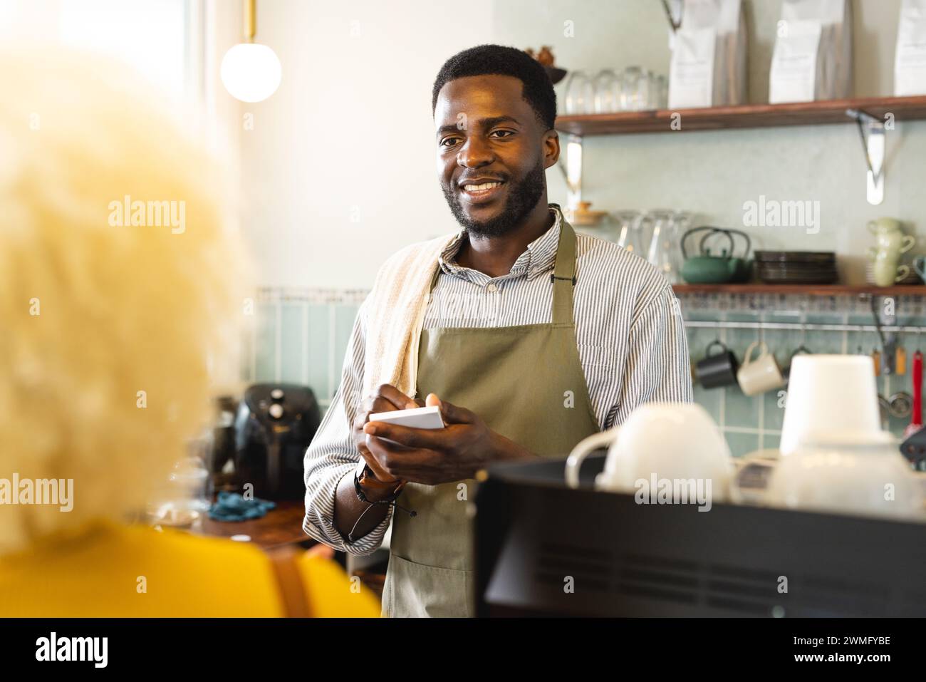 African American barista serves coffee at a cozy cafe. His friendly ...