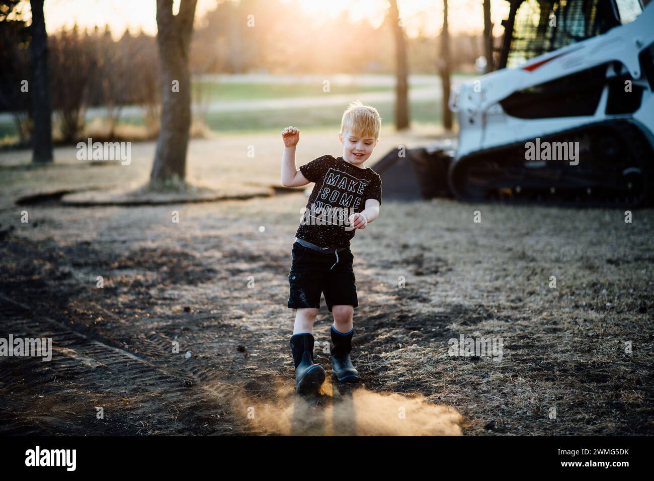 Wide view of young boy kicking up dirt in yard in front of skidsteer ...