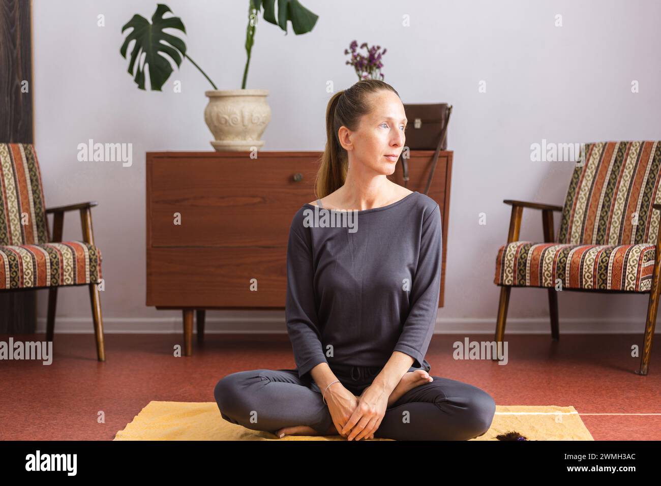 A young adult woman sits in the lotus position in a room and looks out ...
