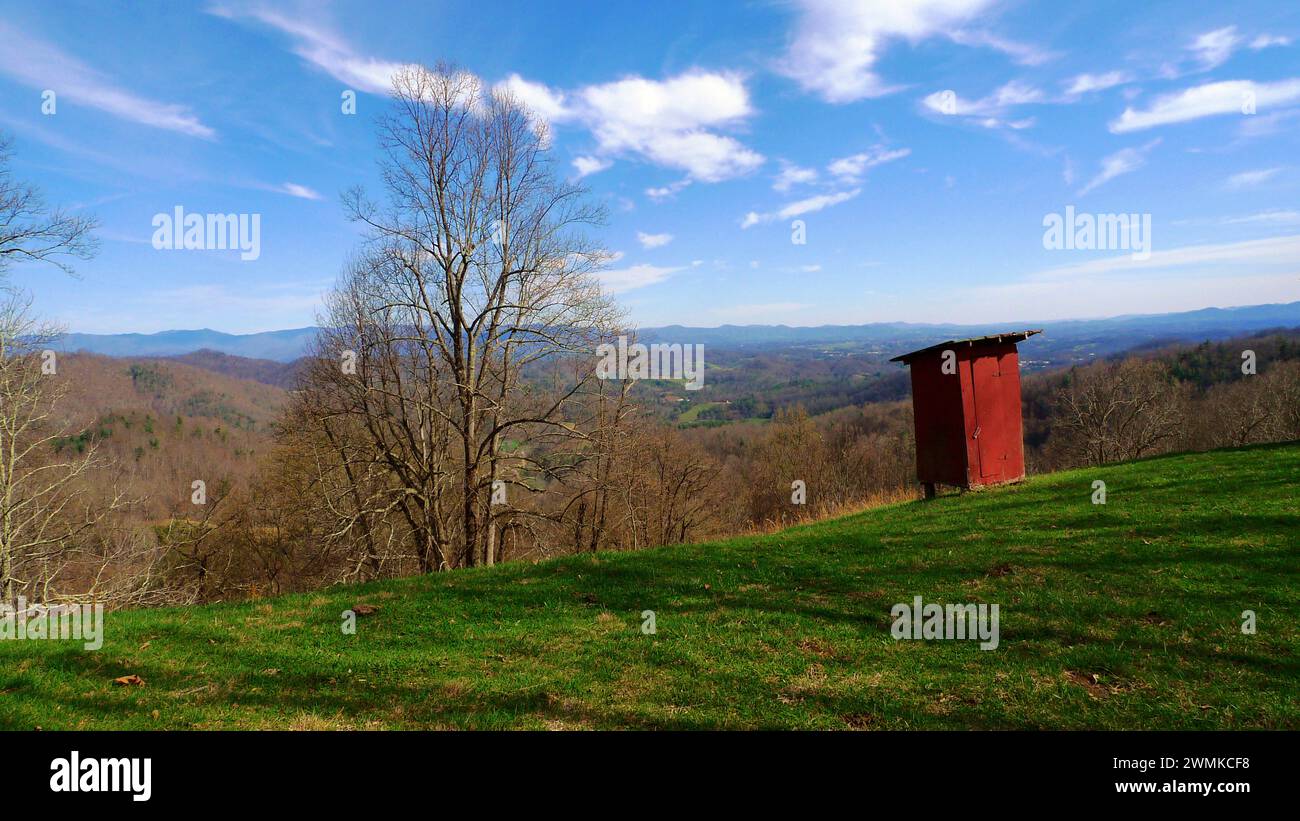 Red outhouse sits on a grass field at the top of a hill, with a view to ...