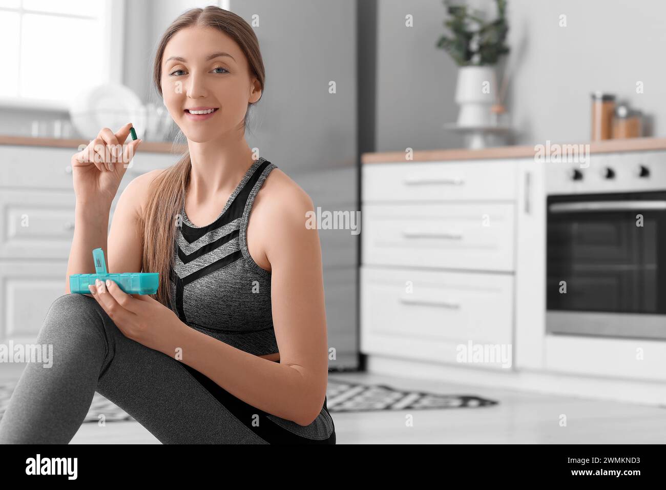 Sporty young woman with container of vitamin supplements in kitchen ...