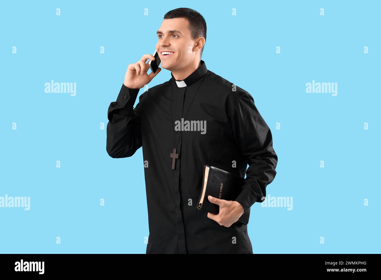 Young priest with Holy Bible talking by mobile phone on blue background ...