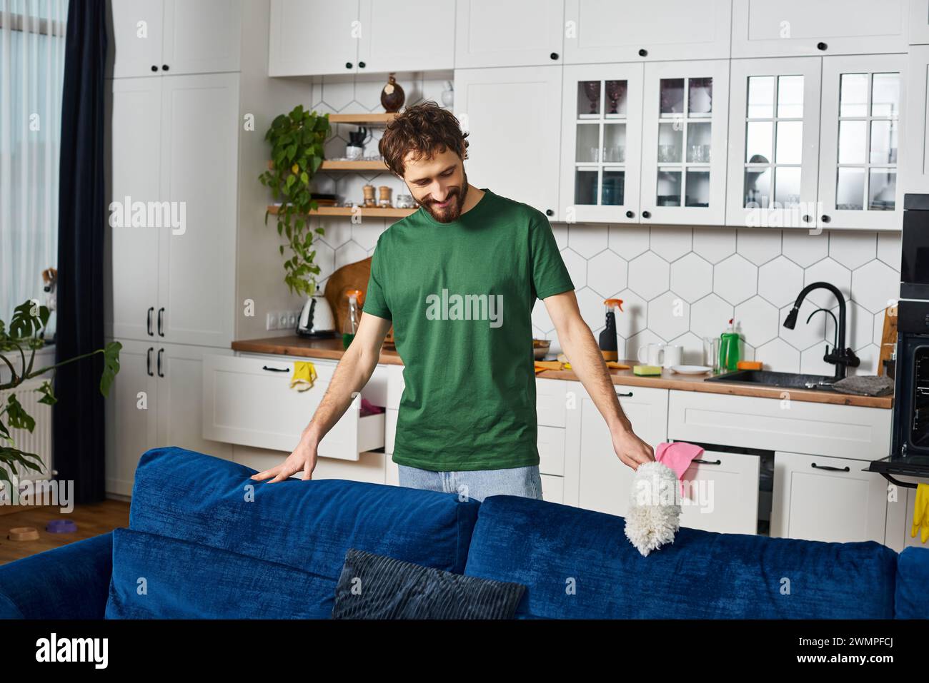 cheerful handsome man in cozy homewear cleaning sofa with duster and ...