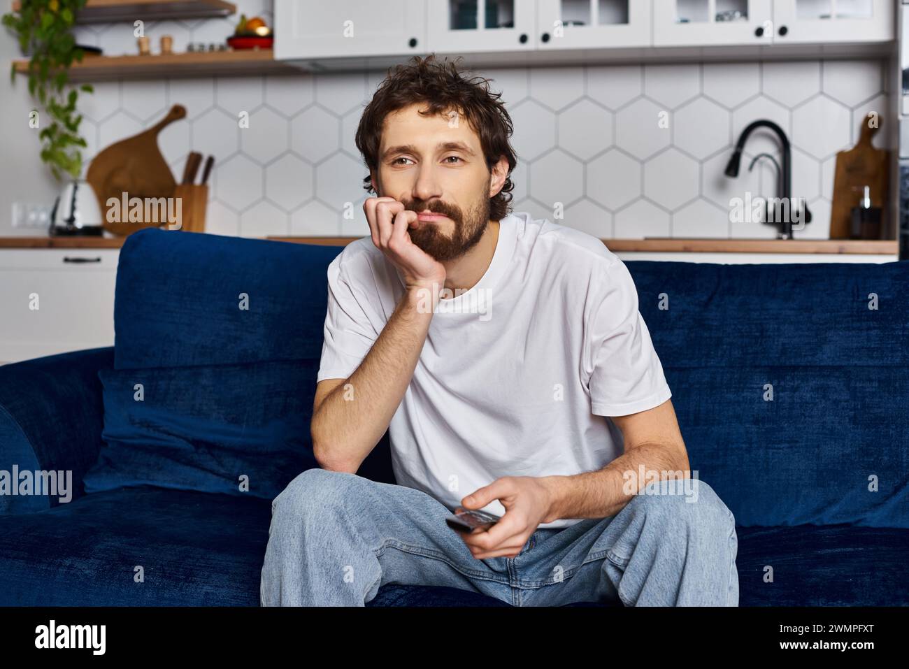 bored handsome man in cozy homewear sitting on sofa with remote control ...