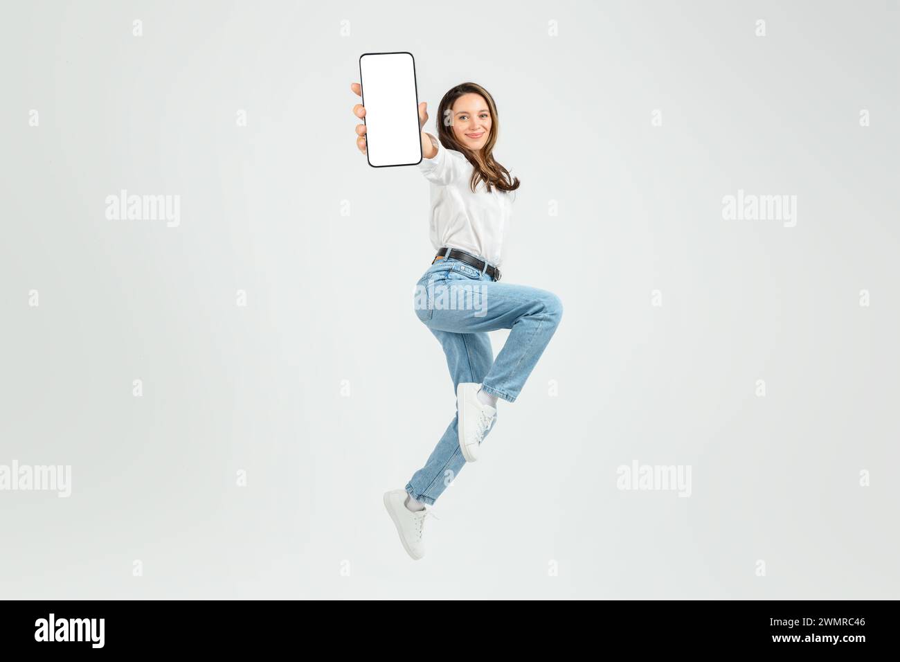 Dynamic young woman in mid-air jump, joyfully presenting a smartphone ...