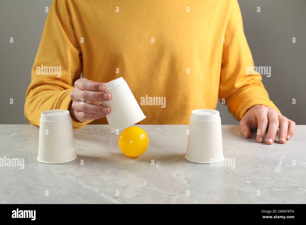 Shell game. Man showing ball under cup at light marble table, closeup ...