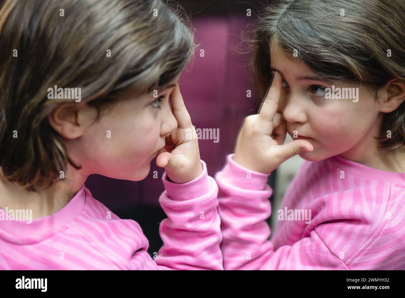 A young girl in a pink top contemplates her reflection in a mirror ...